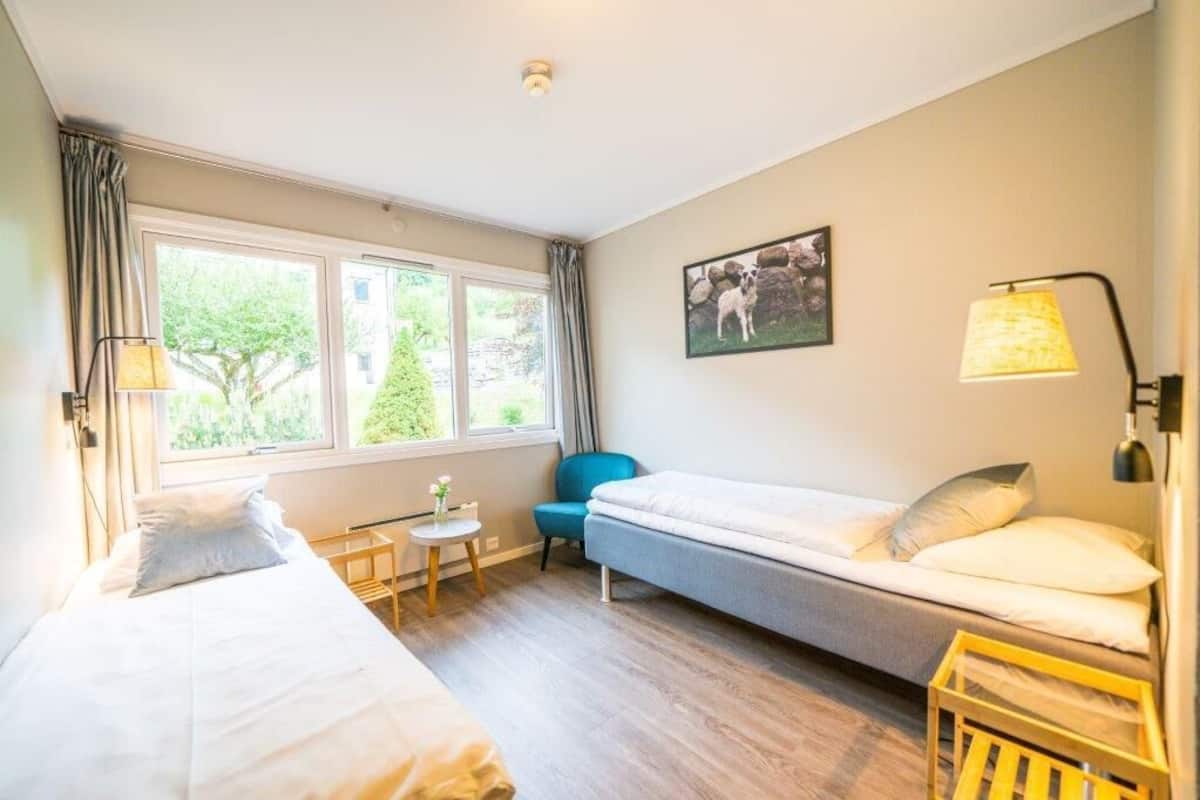 Quarto com duas camas, uma mesinha, uma cadeira e uma janela com cortinas.