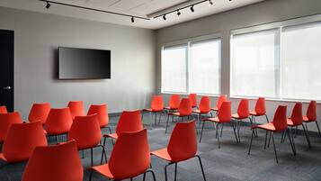 Uma sala de conferências com fileiras de cadeiras laranja, uma TV de tela plana na parede e grandes janelas que oferecem luz natural.