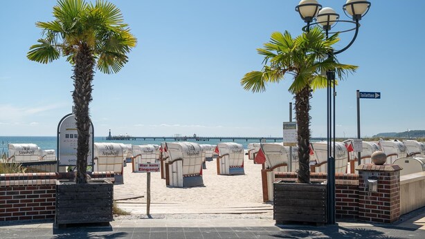 Entrada para a praia com palmeiras, um sinal indicando banheiros, e vista para o mar e um píer ao fundo.