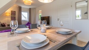 Uma sala de jantar com uma mesa posta para quatro pessoas, incluindo pratos, talheres e copos de vinho. Há duas cadeiras, um sofá e uma televisão ao fundo.