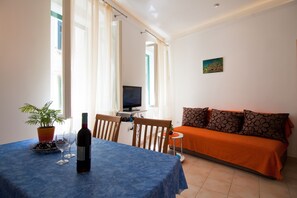 Uma sala de estar com uma mesa azul, um sofá com almofadas estampadas, uma televisão e um vaso de planta.