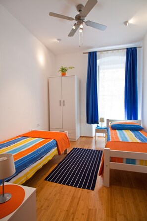 Quarto com duas camas de solteiro, um guarda-roupa, ventoinha no teto e janela com cortinas.