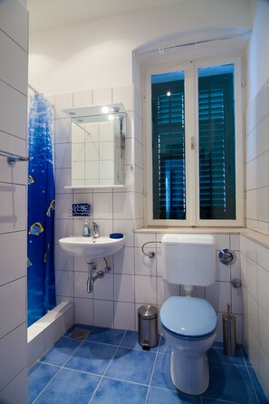 Casa de banho com lavatório branco, sanita, cortina de chuveiro azul e janela com persianas.