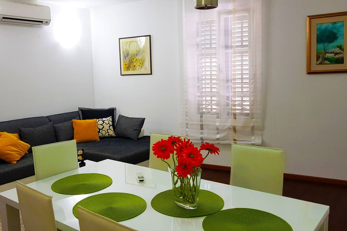 Uma sala de estar moderna com um sofá cinzento, uma mesa de jantar branca com jogos americanos verdes e um vaso com flores vermelhas.