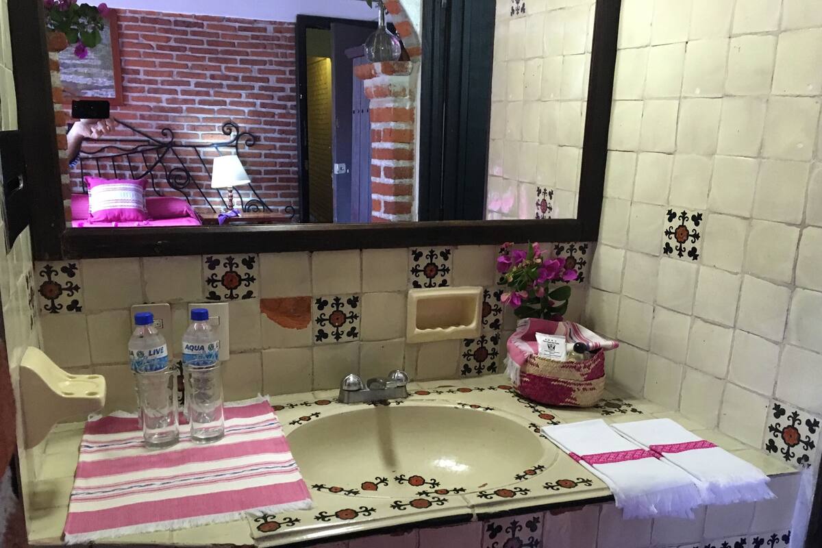 Casa de banho com pia, duas garrafas de água, espelho e cesto decorativo.