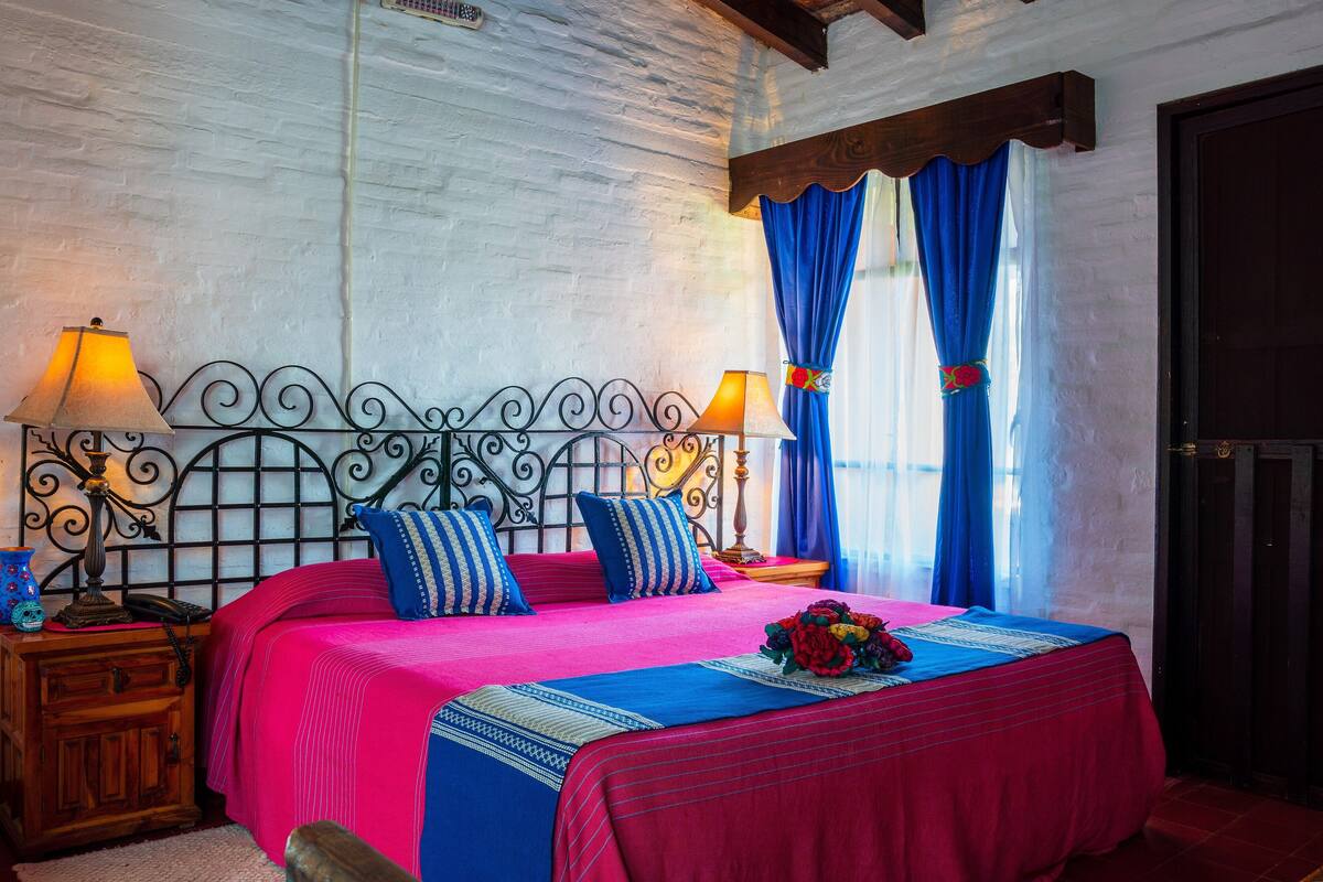 Quarto com uma cama, duas mesas de cabeceira, uma luminária e uma cortina.