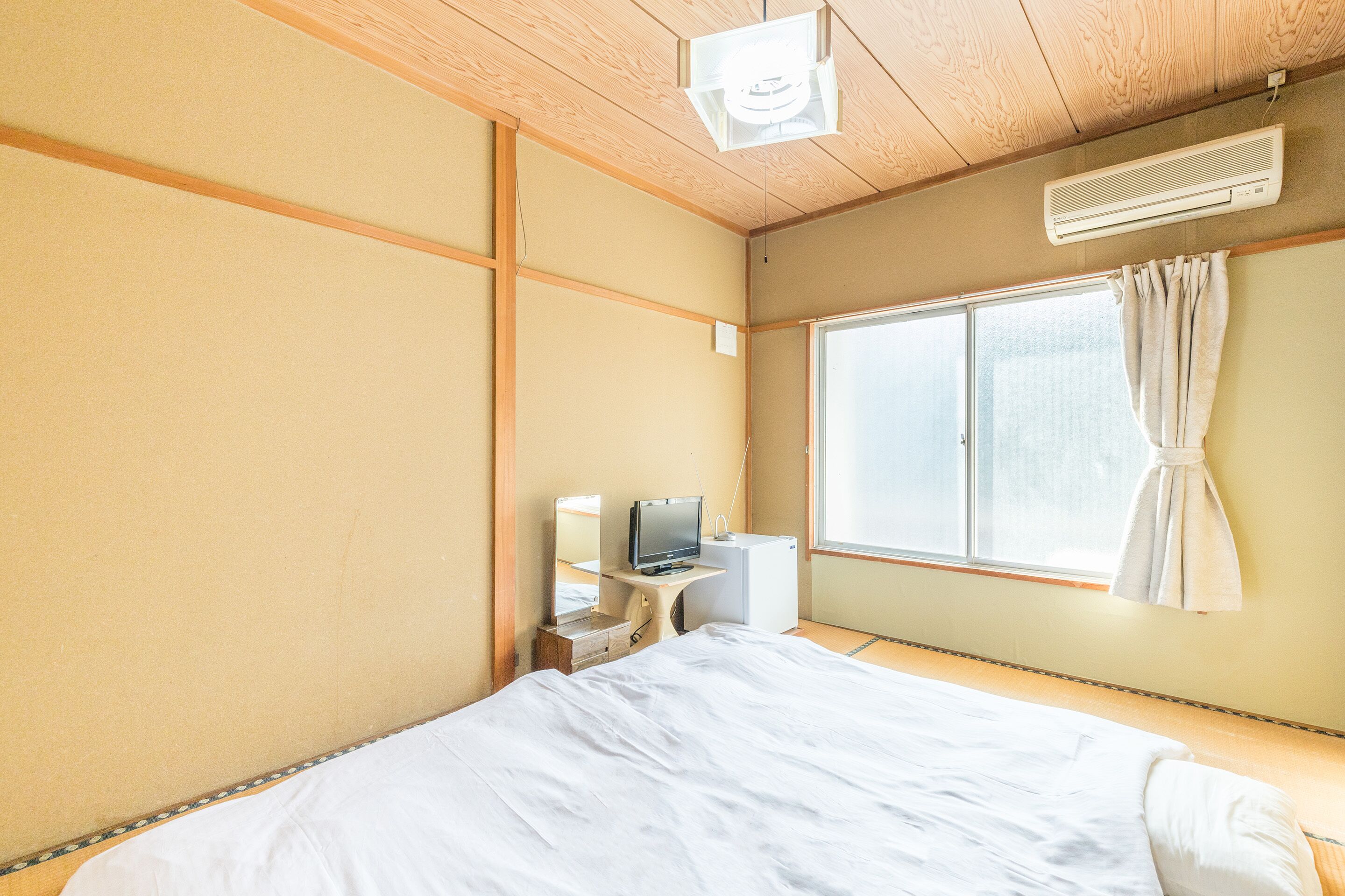 Quarto de hotel com cama, escrivaninha com computador, janela com cortinas e ar condicionado.