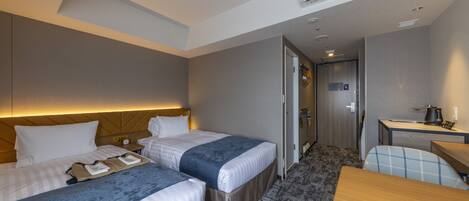 Quarto de hotel com duas camas, uma escrivaninha, uma cadeira e uma cozinha compacta.