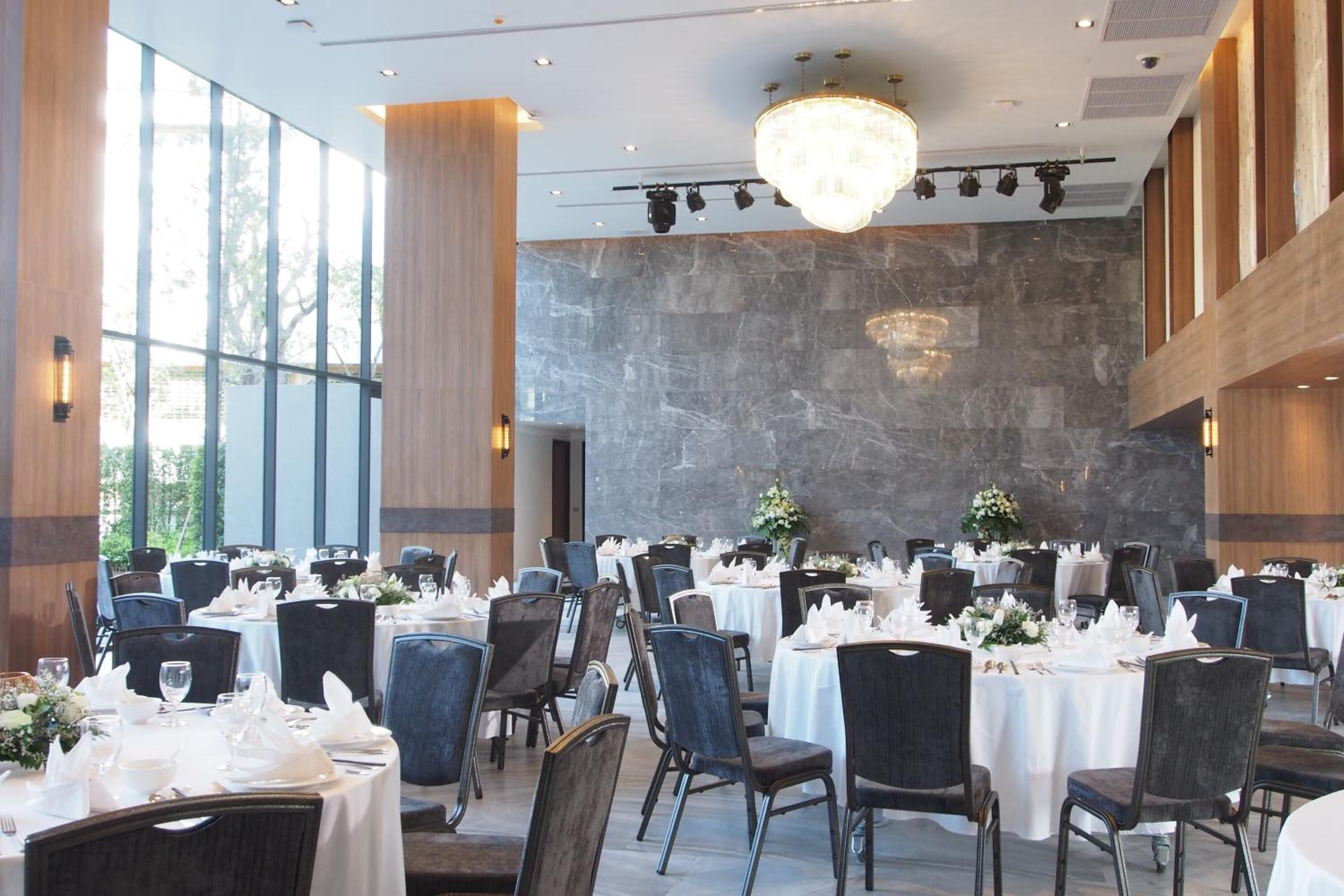 Uma sala de banquetes com mesas redondas preparadas para refeições, janelas amplas e uma parede de pedra.