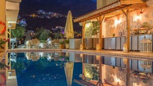 Uma área à beira da piscina com vista clara para uma montanha ao fundo, um espaço coberto com área de estar e um quadro de menu, e um reflexo na água.