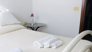 Uma cama bem arrumada com toalhas brancas dobradas, uma mesa redonda pequena com um abajur, e uma porta à direita.