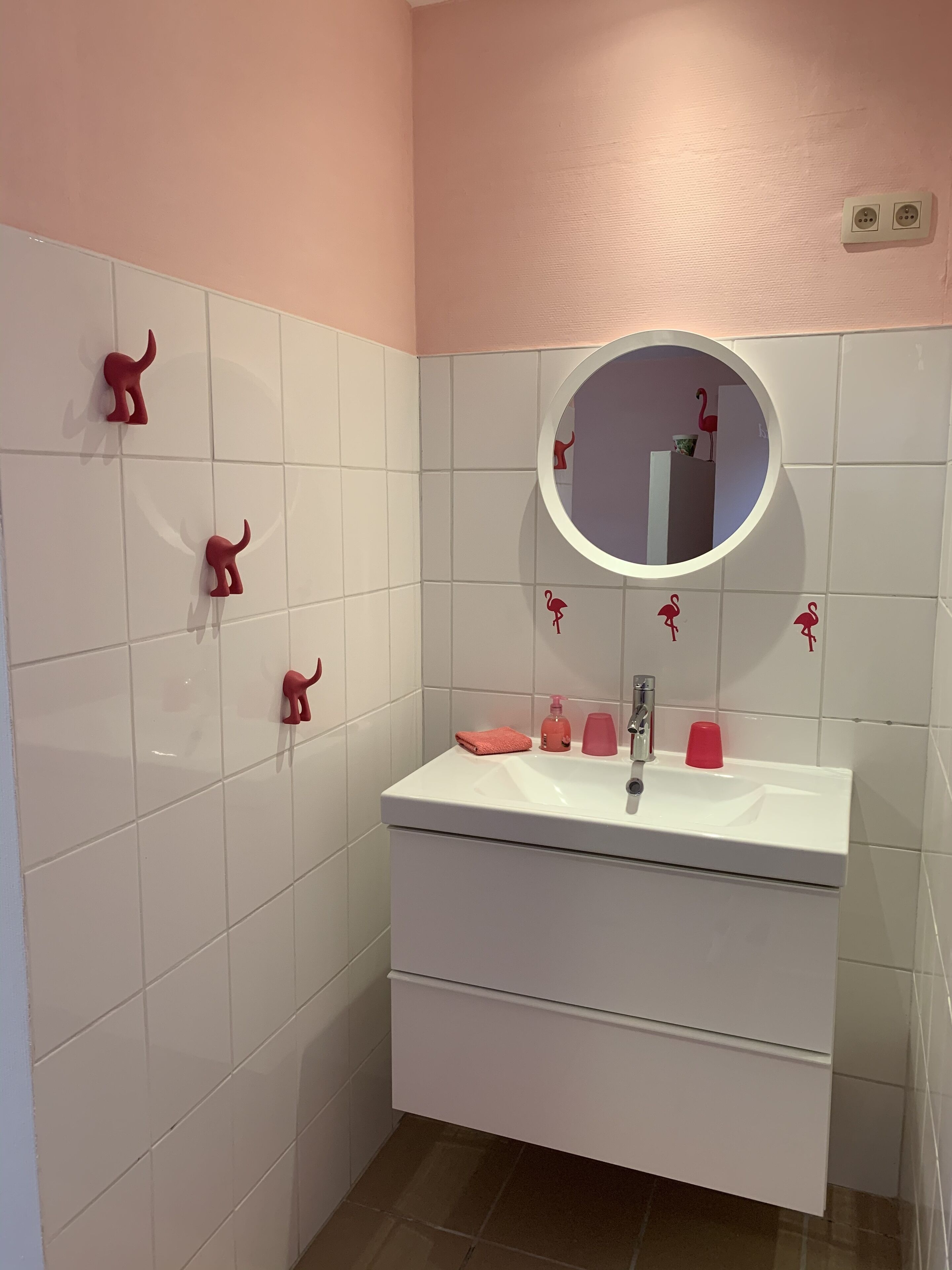 Casa de banho com paredes revestidas a mosaico branco, uma sanita branca com um espelho em cima e três porta-toalhas em forma de flamingo vermelho.