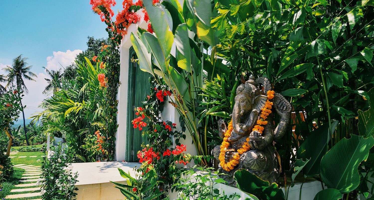 Uma estátua de divindade com forma de elefante, decorada com guirlandas, cercada por vegetação tropical exuberante e um caminho que leva a um edifício com persianas verdes.