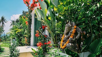 Uma estátua de divindade com forma de elefante, decorada com guirlandas, cercada por vegetação tropical exuberante e um caminho que leva a um edifício com persianas verdes.