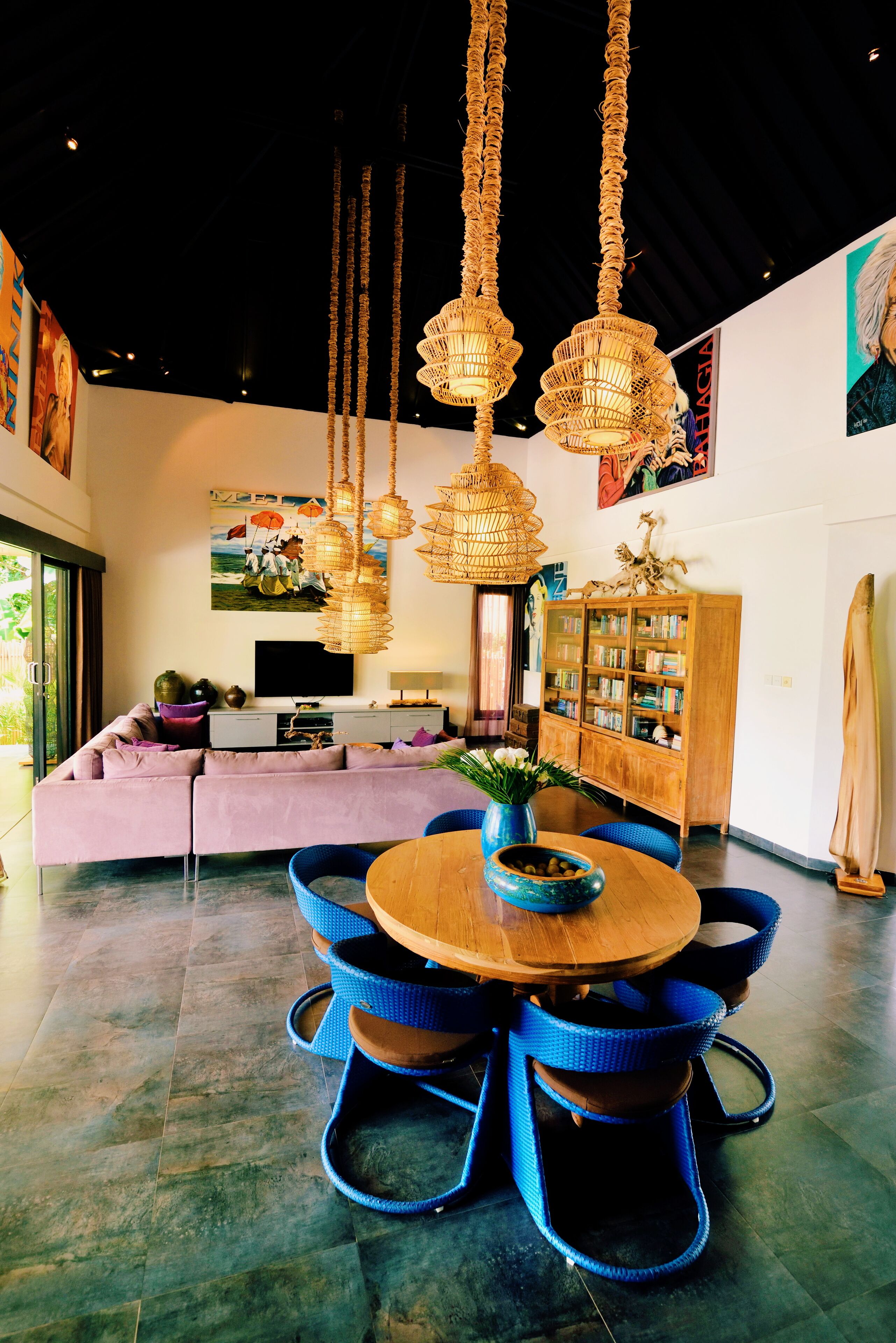 Uma sala de estar moderna com uma mesa de madeira redonda, cadeiras azuis e luminárias de vime suspensas.