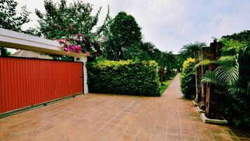 Uma trilha pavimentada conduz a um portão vermelho, cercado por vegetação exuberante e diversas plantas.
