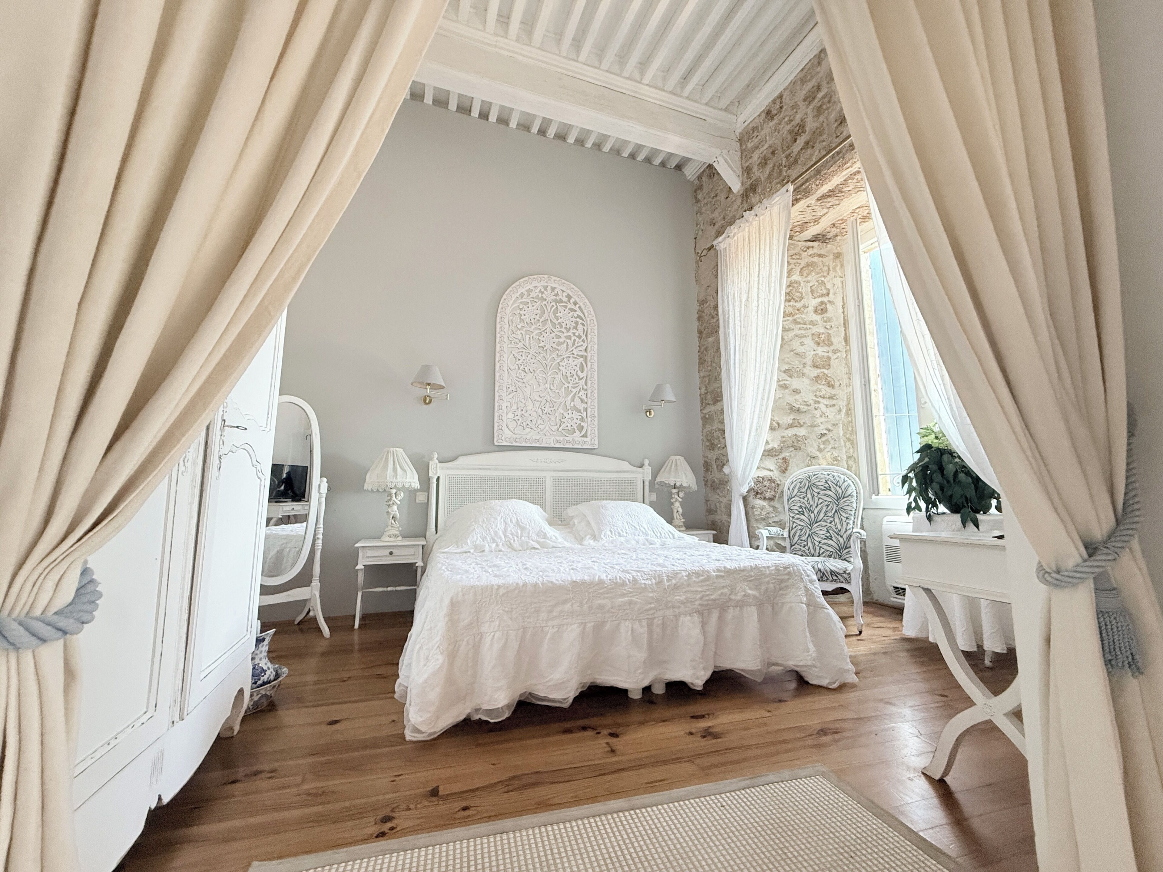 Quarto com cama, mesa de cabeceira, cadeira, abajur, espelho e janela com cortinas.