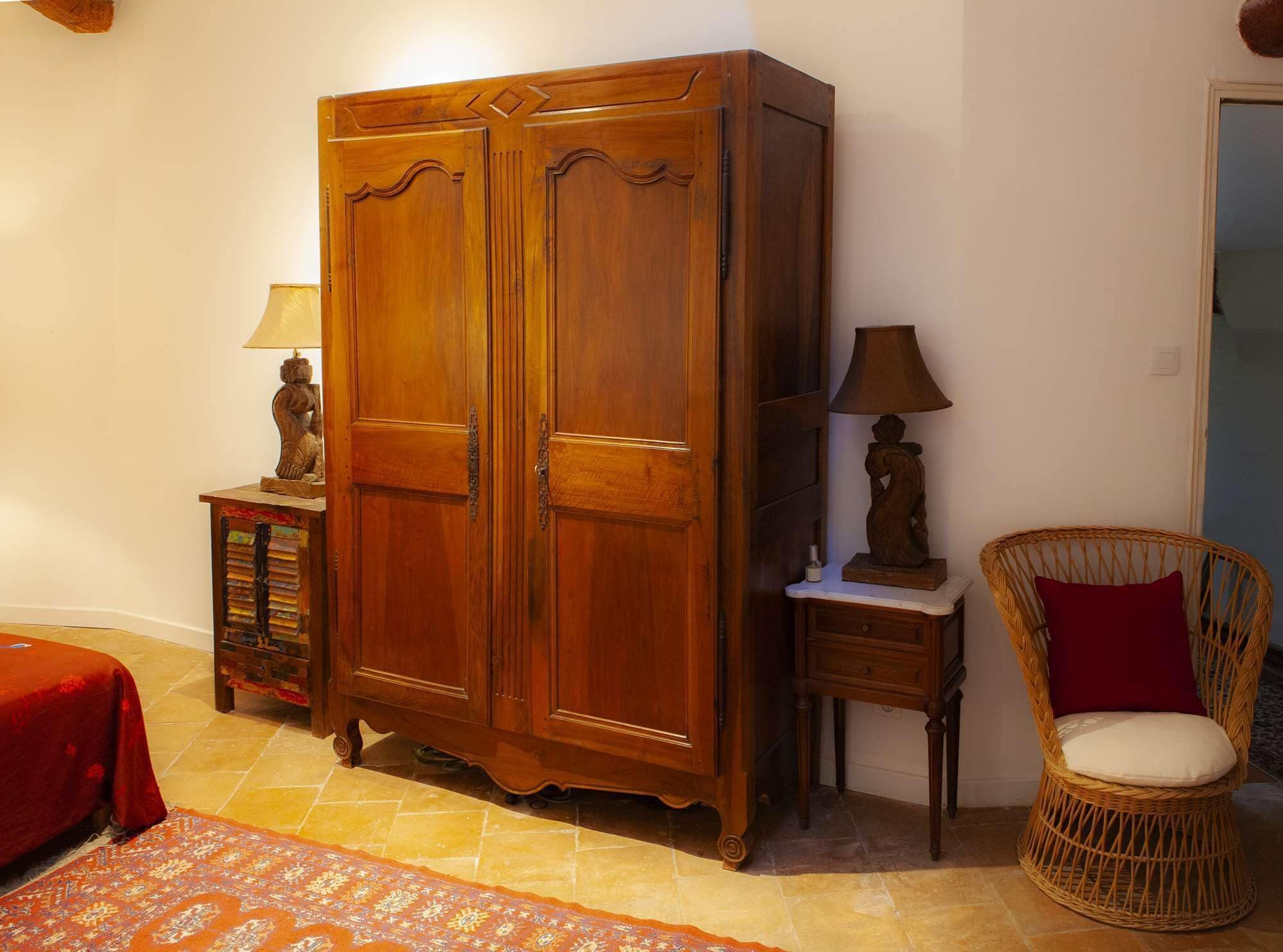 Quarto com um guarda-roupa de madeira grande, uma pequena mesa de cabeceira com luminária, uma cadeira de vime com almofada e um cobreleiro vermelho.
