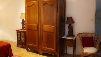 Quarto com um guarda-roupa de madeira grande, uma pequena mesa de cabeceira com luminária, uma cadeira de vime com almofada e um cobreleiro vermelho.