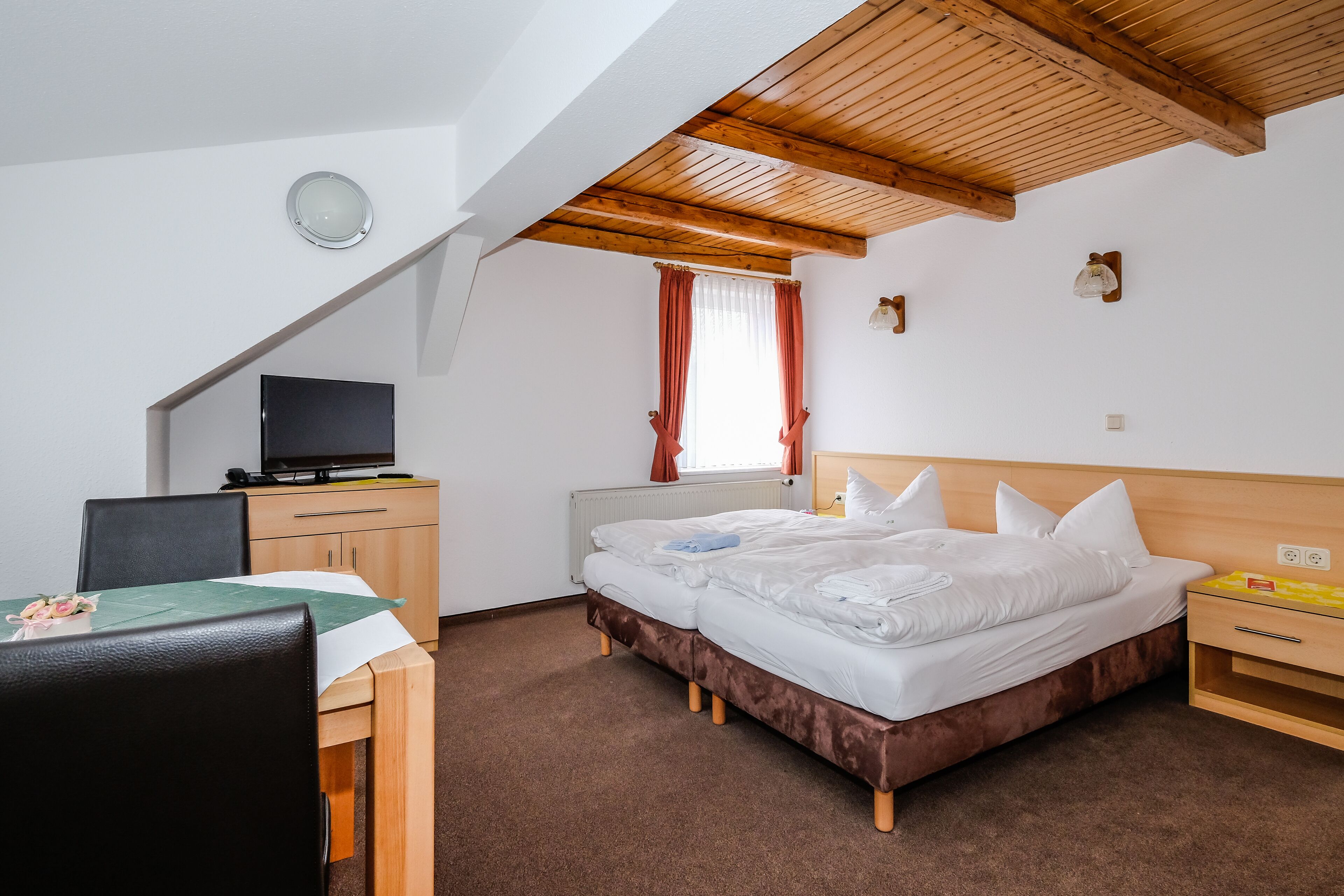 Quarto de hotel com teto de madeira, cama com lençóis brancos, escrivaninha com computador e janela com cortinas vermelhas.