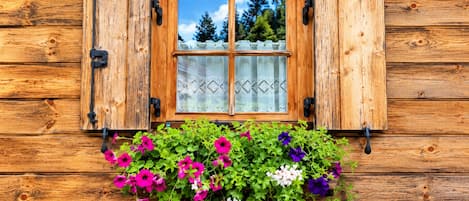 Uma cabana de madeira com janela, cortina decorativa e floreira com diversas flores.
