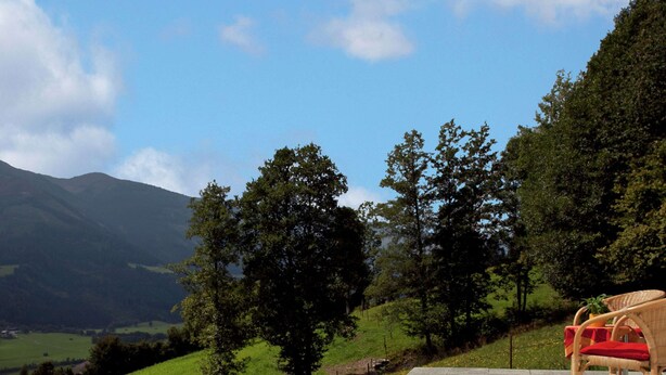 Um terraço com cadeira e mesa de vime, com vista para uma encosta verde e montanhas.