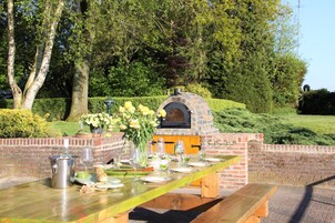 Área de refeições ao ar livre com forno de tijolos, mesa e cadeiras de madeira, cercada por vegetação.