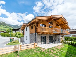 Uma casa moderna de dois andares com varanda de madeira, base de pedra e um pátio pavimentado.