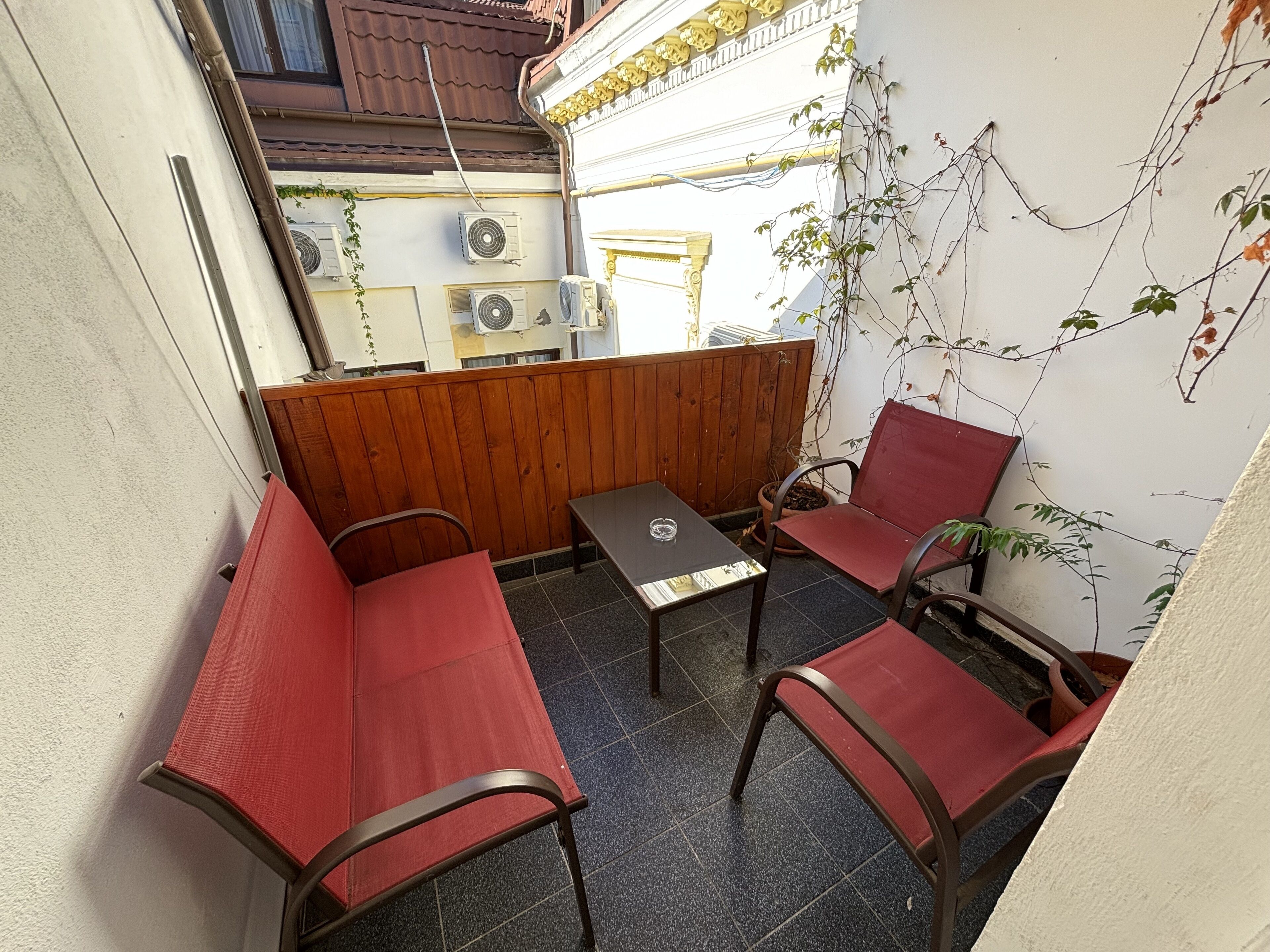 Uma pequena área externa com bancos de madeira e uma mesa, cercada por plantas em vasos e uma parede branca.