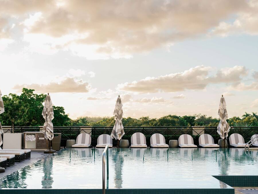 Uma área à beira da piscina com espreguiçadeiras e guarda-sóis, céu claro e vista para árvores ao fundo.