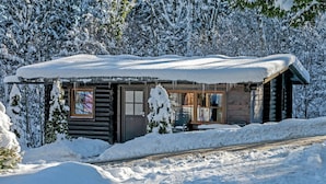 Uma cabana de toras com telhado coberto de neve, rodeada por árvores.