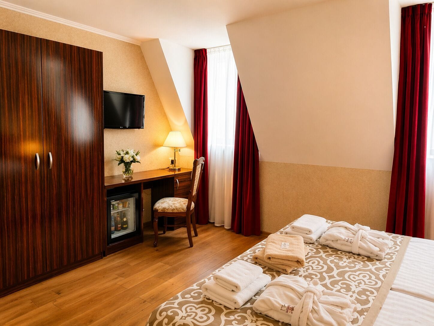 Quarto de hotel com guarda-roupa de madeira, escrivaninha com candeeiro, cadeira, cama com toalhas e televisão na parede.