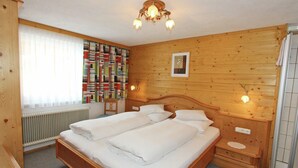Uma cama de casal com lençóis brancos, cabeceira de madeira, mesa de cabeceira, pequena mesa de apoio, radiador, janela com cortinas e luminária fixada na parede.