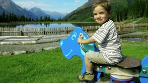 Criança sentada em um carrinho de pedal azul e verde em um parque com um lago e montanhas ao fundo.