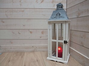 Um candeeiro branco com uma vela no interior, colocado sobre um soalho de madeira, em frente a uma parede de madeira.