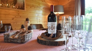 Interior de uma cabana de madeira com uma mesa posta para refeição, incluindo uma garrafa de vinho, copos, pinhas e uma cesta de vime.