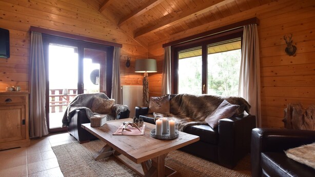 Interior acolhedor de uma cabana, com teto de madeira, um sofá de couro, uma mesa de madeira com velas e grandes janelas com cortinas.