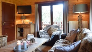 Interior acolhedor de uma cabana, com teto de madeira, televisão na parede, área de jantar com mesa e velas, e um sofá com manta.
