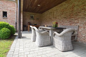 Um pátio externo com parede de tijolos, uma mesa, cadeiras de vime e um banco.