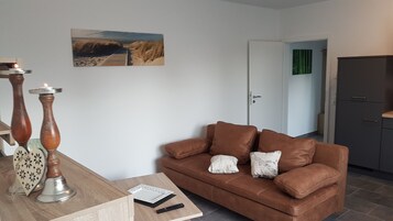 Uma sala de estar com um sofá de couro marrom, uma mesa de centro de madeira, dois vasos decorativos e um quadro com uma paisagem de praia na parede.