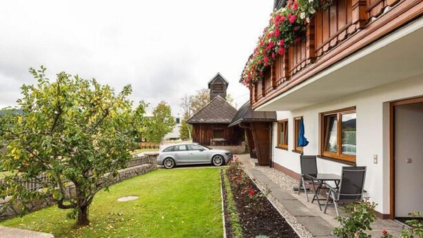 Uma casa com um carro estacionado na entrada, um pequeno jardim com uma árvore e um cercado de madeira.