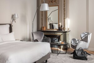 Quarto de hotel com cama, duas poltronas, uma mesa redonda, lareira e espelho.