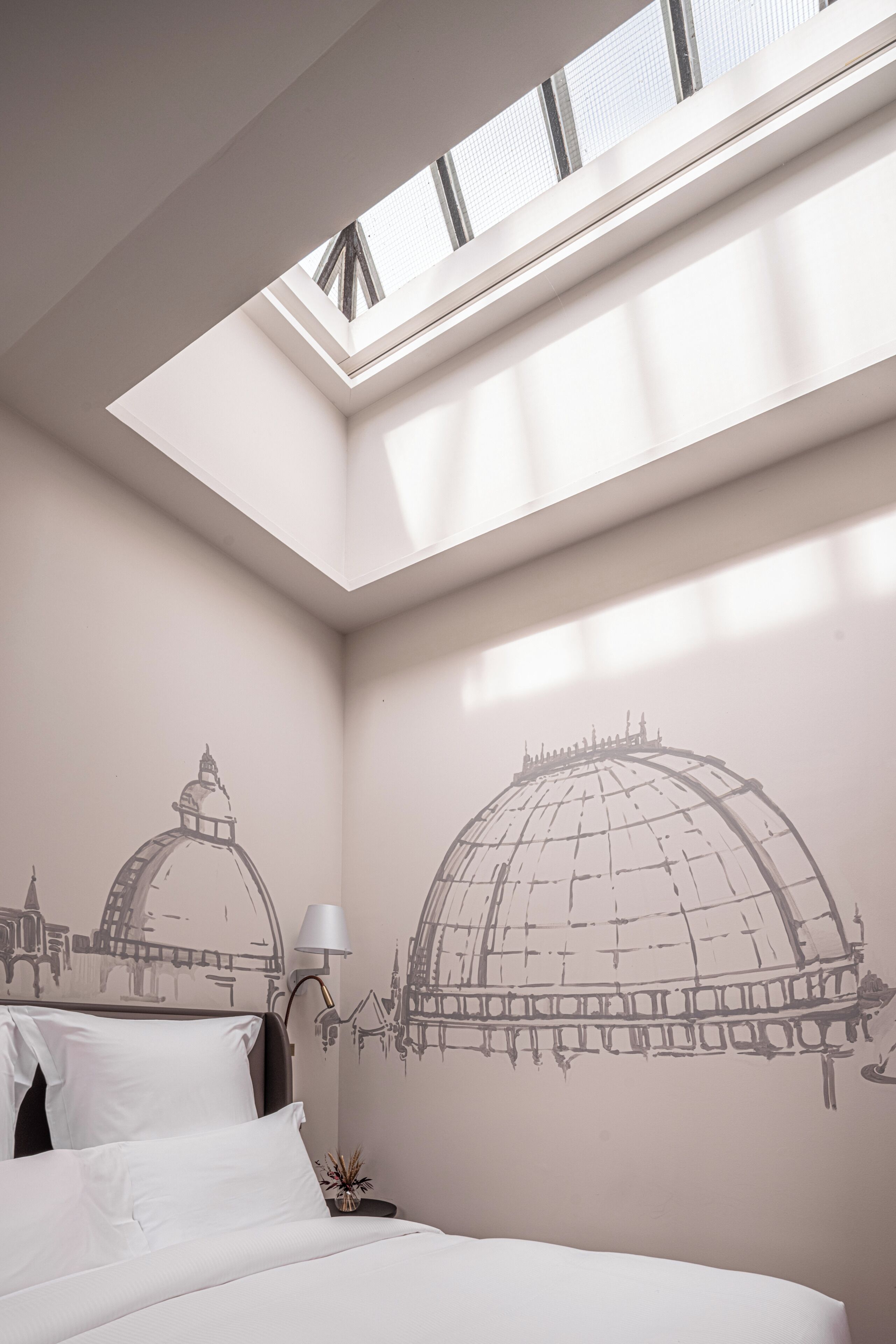 Quarto com claraboia, cama com roupa de cama branca, mesa de cabeceira com abajur e um mural na parede com cúpulas arquitetônicas.