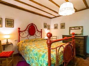 Quarto com estrutura de cama de madeira, cabeceira vermelha, colcha estampada, mesa de cabeceira com abajur, cadeira, cômoda e quadros nas paredes.