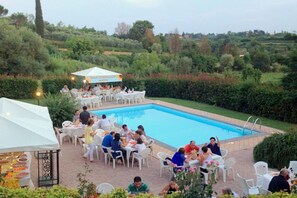 Uma área de piscina ao ar livre com mesas e cadeiras para refeições, cercada por vegetação e um céu azul límpido.