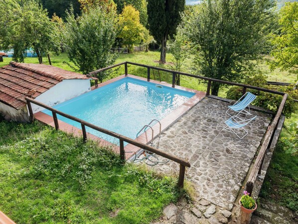 Piscina retangular com deck de pedra e guarda-corpo de madeira. Há duas espreguiçadeiras azuis listradas e um pequeno edifício branco com telhado em azulejo.
