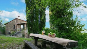 Um edifício rústico de pedra com uma escadaria de pedra que leva a uma porta de madeira. Um banco de madeira com uma mesa está disposto à frente, coberto por um tecido, e há cestas de maçãs e garrafas sobre a mesa.