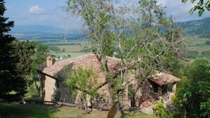 Um edifício rústico de pedra com telhado de azulejos, rodeado por vegetação exuberante e uma paisagem montanhosa ao fundo.