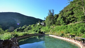 Uma piscina natural rodeada por rochas e vegetação, com uma casa ao fundo.