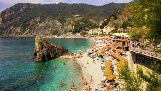 Uma praia costeira com águas turquesa cristalinas, falésias rochosas e uma fileira de edifícios à beira-mar com guarda-sóis.
